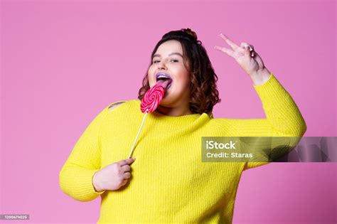 밝은 플러스 크기 모델 큰 롤리팝 분홍색 배경에 노란색 점퍼에 뚱뚱한 여자 건강에 좋지 않은 음식에 대한 스톡 사진 및 기타