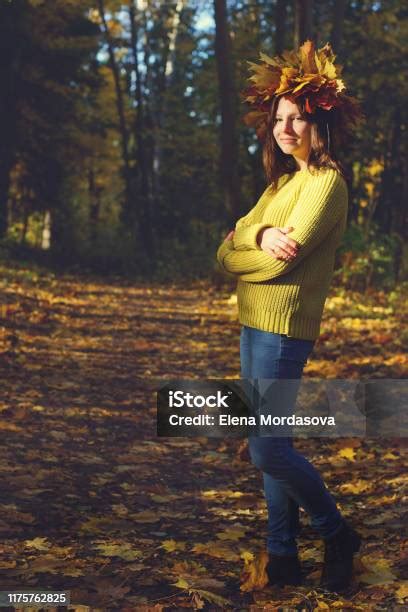 노란 스웨터와 청바지를 입은 어린 갈색 머리 소녀가 가을의 상징인 단풍나무 화환을 머리에 머리에 대고 가을 공원의 길에 서 있습니다 가을에 대한 스톡 사진 및 기타 이미지