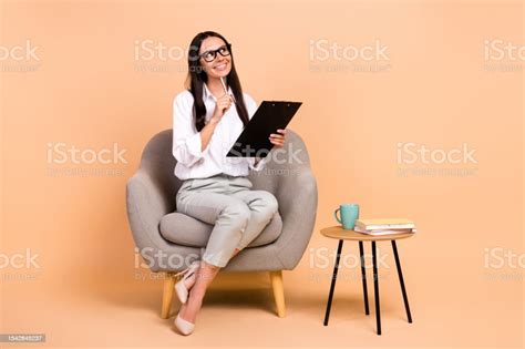 재미 있고 꿈꾸는 아가씨의 전체 길이 사진은 빈 공간을 찾고 있는 문서를 쓰는 흰색 셔츠 앉아있는 의자 격리 된 베이지 색