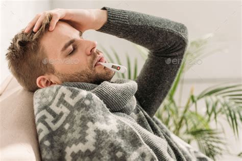 Sick Man Wrapped In Blanket Sitting On Sofa With Closed Eyes And