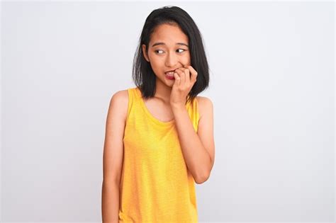 노란 캐주얼 티셔츠를 입은 젊은 중국 여성은 손톱을 물고 손톱을 물고 손에 스트레스와 긴장 찾고 고립 된 흰색 배경 위에 서있다 불안 문제 T 셔츠에 대한 스톡 사진 및