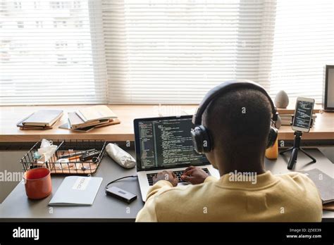 Rear View Of Black Programmer In Wireless Headphones Sitting At Desk With Hard Drive And Using