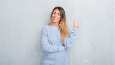 젊은 성인 여자 승리 표시를 하 고 카메라에 윙크 하는 행복 한 얼굴로 웃 고 겨울 옷을 입고 회색 그런 지 벽에 번호 2입니다