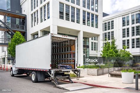 개방형 뒷문과 리프트가 있는 박스 트레일러가 있는 중간 장비 세미 트럭의 재배치 전문가는 다층 사무실 및 아파트 건물이 있는 도시 거리에 서 있는 고객에게 배송합니다 0명에