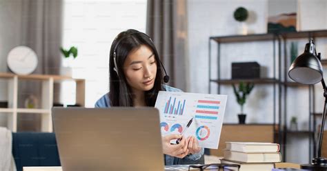 집에서 일하는 동안 노트북에서 화상 회의를 하는 헤드셋을 쓴 진지한 아시아 여성 매력적인 한국 여학생이 책상에 앉아 노트북을 통해 동료들에게 그래프와 차트를 보여주고 있다