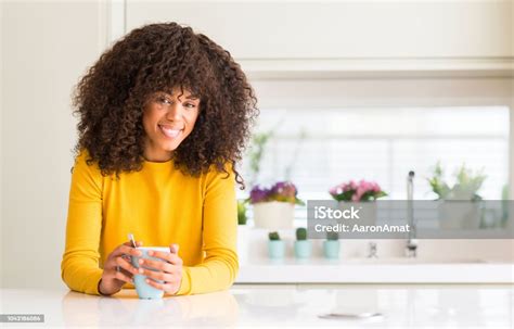 아름 다운 아프리카계 미국인 여 자가 서 이빨을 보여주는 자신감 미소로 웃 고 행복 한 얼굴로 집에서 커피 한 잔을 들고 건강한