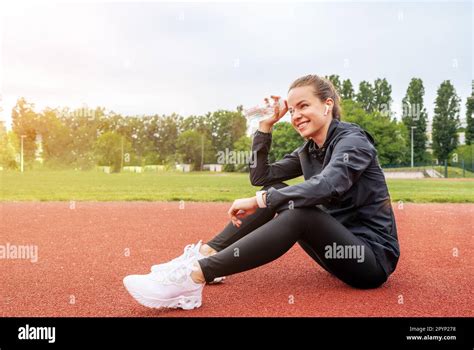 Athletic Young Woman Runner Sitting On Running Track Resting After Training And Smiling Stock