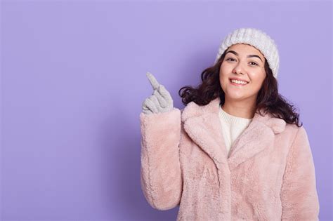 행복한 표정으로 사랑스러운 갈색 머리 여성 핑크 모피 코트 모자와 장갑을 착용 라일락 배경에 대한 스탠드 프로모션을위한 빈 공간을 보여줍니다 20 24세에 대한 스톡 사진