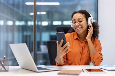 오렌지색 셔츠를 입은 여자가 사무실 환경에서 노트북과 노트북을 가진 책상에서 스마트 폰과 헤드폰을 사용하면서 웃고 있습니다 Brand Name Smart Phone에 대한