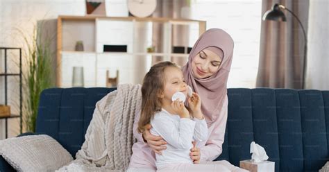 사랑스러운 돌보는 무슬림 엄마 집에서 파란색 소파에 앉아 무릎에 어린 딸을 안고 있습니다 귀여운 행복한 아이는 위생 절차를 만들고 면봉으로 얼굴을 닦습니다 사진