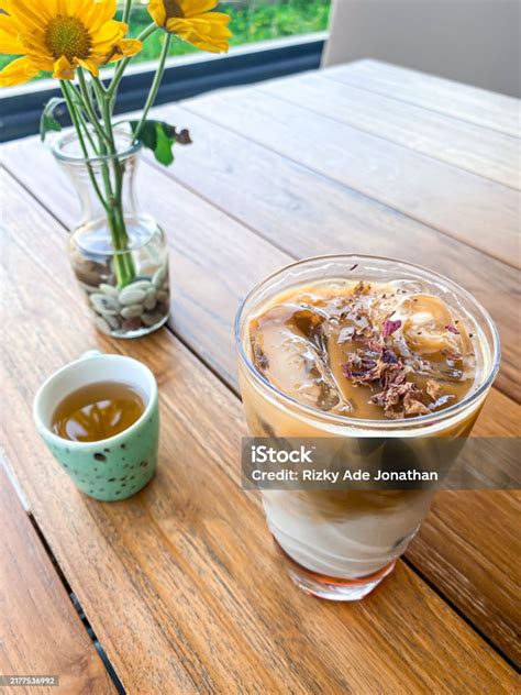 투명한 유리잔에 말린 장미 꽃잎을 뿌린 아이스 카푸치노 커피 나무 테이블 위에 액상 설탕 컵과 꽃병이 놓여 있습니다 머그잔에 담긴 카페 라떼 또는 커피 우유 음료 0명에