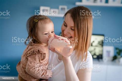 작은 백인 아기 소녀가 집에서 딸을 안고있는 어머니의 손에 병에서 물을 마시는 어린 시절의 발달과 가족 개념 물에 대한 스톡