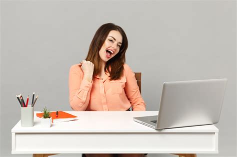 파스텔 옷에 젊은 즐거운 여자는 예 앉아 회색 배경에 고립 된 Pc 노트북과 책상에서 작업을 말하고 위 너 제스처를 하 고