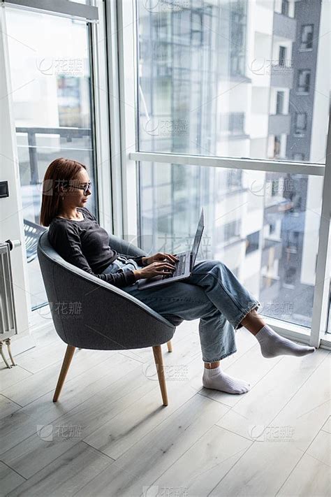 一个微笑的年轻女子坐在靠近窗户的现代椅子上，在家里灯光舒适的房间里。她在轻松的氛围中使用笔记本电脑工素材 花瓣网