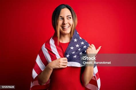 젊은 아름다운 금발의 애국적인 여자 와 푸른 눈을 입고 미국 국기 를 가리키고 행복한 얼굴 미소와 함께 측면에 엄지 손가락으로 보여주는 가리키기에 대한 스톡 사진 및 기타