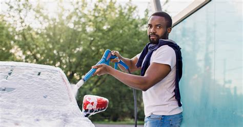 고압 제트를 사용하여 비누 거품으로 자동차의 실외 청소 잘 생긴 수염을 기른 아프리카 청년이 야외에서 셀프 세척 서비스에서 거품으로 고압으로 빨간 차를 씻고 있습니다