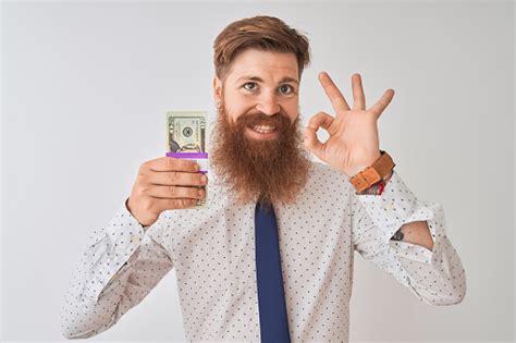 손가락으로 확인 기호를하고 고립 된 흰색 배경 위에 서 있는 달러를 들고 젊은 빨간 머리 아일랜드어 사업가 우수한 기호 개념에