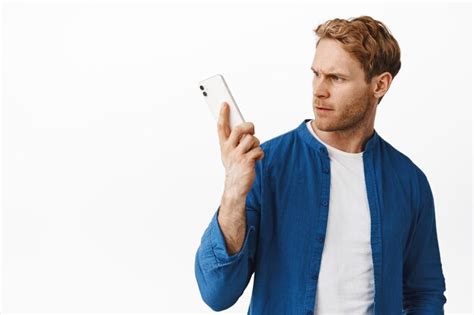 Портрет рыжего парня выглядит смущенным на смартфоне, слышит что-то ...