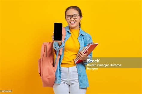 배낭으로 데님 옷을 입고 책을 들고 노란색 배경에 고립 된 빈 화면 휴대 전화를 보여주는 젊은 아시아 여성 학생의 초상화 25