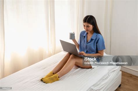 아름다운 얼굴을 한 젊은 아시아 여성 파란 셔츠를 입은 긴 머리가 집에서 흰색 침실 침대에 앉아 커피 한 잔과 노트북으로 화상