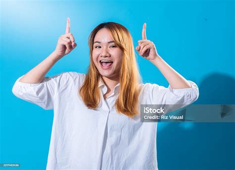 긴 생머리를 한 아시아 여성이 부드러운 스튜디오 섀도우와 쾌활한 분위기의 생생한 파란색 배경에 흰색 셔츠를 입고 두 검지 손가락을 위로 들어 올리면서 밝게 미소 짓고