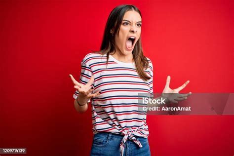 젊은 아름 다운 갈색 머리 여자 캐주얼 줄무늬 티셔츠 를 입고 빨간 배경 미친 미친 위에 서 미친 소리와 공격적인 표정과 팔을 제기 고함 좌절 개념입니다 T 셔츠에 대한