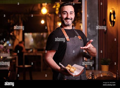 Waiter Looking At The Camera And Smiling While Showing Plate With Food In A Restaurant Stock