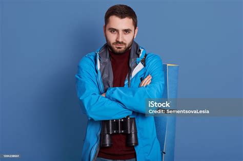 접힌 손으로 파란색 배경 위에 고립 된 수염을 가진 잘 생긴 남자의 수평 샷 매트와 쌍안경을 가진 여행자 재킷을 입고 카메라를