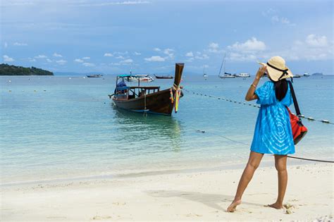 빨간 방수 가방이 달린 파란색 드레스를 입은 젊은 여성 흰색 모자에 손잡이 하얀 해변에 서서 바다의 아름다움과 태국 크라비 주 피 피 섬에서 현지 롱 테일 보트를 보고