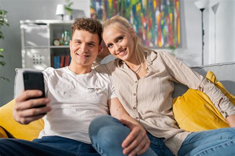 친구의 행복한 부부 그들은 카메라를보고 거실에서 소파에 앉아 소년은 셀카를 찍기 위해 그 앞에 뻗어 전화를 들고 사진 소녀는 웃고있다 2명에 대한 스톡 사진 및 기타 이미지