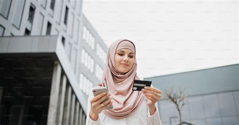 공식적인 옷을 입고 히잡을 쓴 아름다운 무슬림 여성이 신용 카드와 스마트폰으로 온라인 쇼핑을 하고 있다 사무실 건물 근처에 서서 쉬운 지불을 즐기는 젊은 아가씨 사진