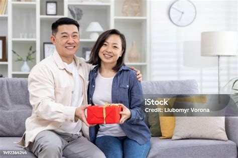 젊은 아시아 커플 남자와 여자가 집에 있는 소파에 앉아 선물 상자를 손에 들고 있다 카메라를 바라보며 웃고 행복해하는 모습 선물에 대한 스톡 사진 및 기타 이미지 Istock