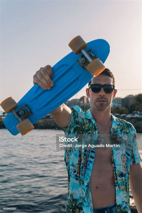 파란색 오픈 셔츠와 검은 색 선글라스를 입은 검은 색 선글라스를 입은 어두운 머리 소년의 수직 이미지로 한 손에 푸른 페니 롱 보드를 들고 배경에 바다가 있습니다 중년