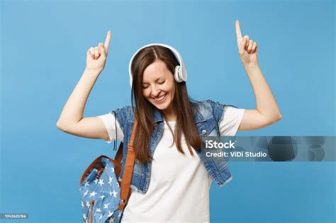젊은 예쁜 여자 학생 배낭 닫힌된 눈 헤드폰 파란색 배경에 고립 된 춤 검지 손가락을 가리키는 하는 음악 들 고등학교에서