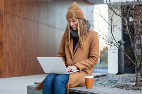 벤치에 앉아 활짝 웃는 젊은 여자는 도시 거리에서 커피를 손에 들고 노트북을 본다 사무실이 아닌 관리자는 랩톱에서 추가 작업에