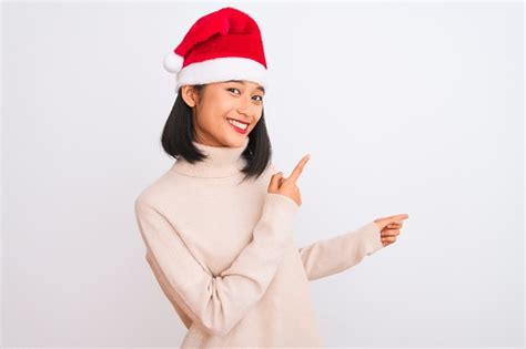 고립 된 흰색 배경 위에 크리스마스 산타 모자를 쓰고 미소를 짓고 두 손과 손가락으로 가리키는 카메라를 바라보는 젊은 아름다운 중국 여성 12월에 대한 스톡 사진 및 기타
