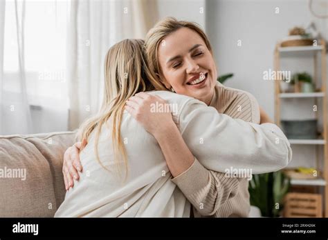 Two Blonde Sisters In Lovely Casual Attire Hugging On Sofa With Closed