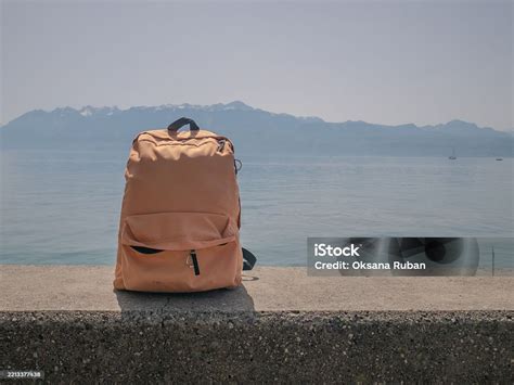 산맥을 배경으로 잔잔한 호수의 콘크리트 제방에 놓여 있는 외로운 베이지색 배낭 솔로 여행과 최소한의 관광이라는 개념 부드러운 대낮 아래 정면 0명에 대한 스톡 사진 및 기타