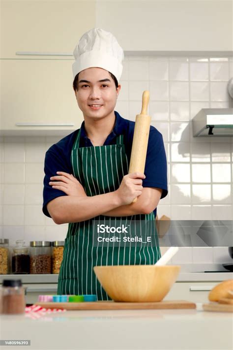 앞치마와 요리사 모자를 쓴 젊은 행복한 미소를 짓고 있는 아시아 남자의 초상화는 나무 롤링 반죽 스틱을 들고 부엌에 팔을 들고