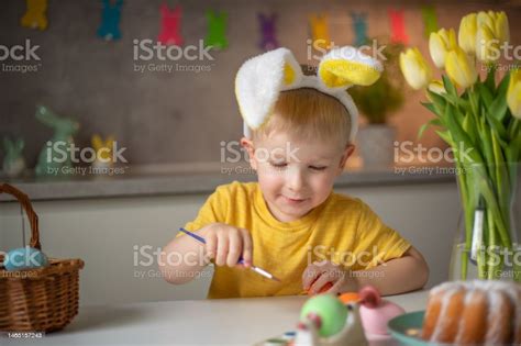 토끼 귀를 쓴 어린 소년은 식탁에 앉아 부활절을 위해 색칠한 부활절 달걀을 준비합니다 행복한 부활절 2 3 살에 대한 스톡 사진