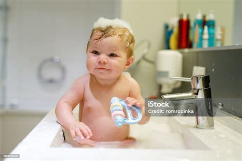 세면대에 목욕을하고 귀여운 사랑스러운 아기는 물 탭을 잡아 큰 파란 눈을 가진 작은 건강한 소녀는 비누 거품을 가지고 놀고 있습니다 가정의 방에 대한 스톡 사진 및 기타