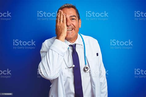한 눈을 손으로 덮고 얼굴에 자신감 있는 미소와 깜짝 감정을 덮고 있는 고립된 파란색 배경 위에 청진기를 착용한 잘생긴 중년 의사 남자 가리기에 대한 스톡 사진 및 기타