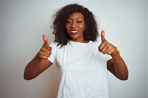 고립 된 흰색 배경 성공 표지판 위에 서있는 티셔츠를 입은 젊은 아프리카 계 미국인 여성은 손으로 긍정적 인 제스처를하고 엄지 손가락으로 미소와 행복을 합니다 쾌활한 표정과