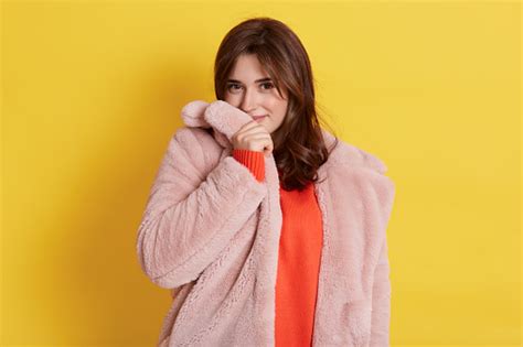 꿈꾸는 신비한 미소를 가진 갈색 머리 소녀는 오렌지 색 스웨터와 옅은 분홍색 모피 코트를 입고 칼라에 손을 잡고 뺨을 숨기고 노란색 벽에 서 있습니다 20 24세에 대한