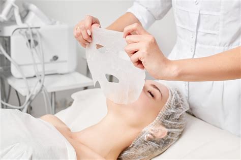 미용사는 얼굴 피부 재수화를 위해 여성 얼굴에 시트 마스크를 바르고 뷰티 살롱에서 시술합니다 25 29세에 대한 스톡 사진 및