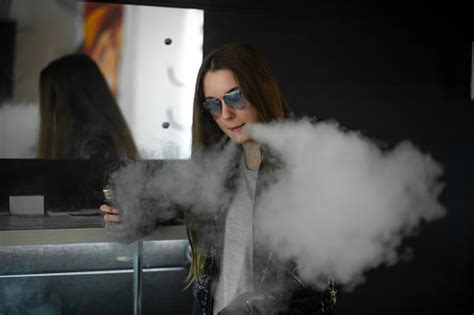 Vape 십대 문제 피부 선글라스를 쓴 어린 귀여운 소녀의 초상화가 거울 맞은편에 전자 담배를 피우고 있습니다 나쁜 습관