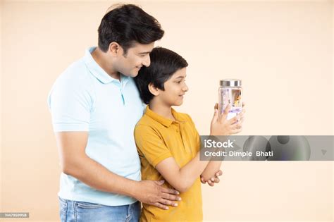 행복한 젊은 인도 아버지와 아들이 베이지색 스튜디오 배경에 고립된 루피 지폐로 가득 찬 유리병을 들고 있다 돈을 절약하고 개념을 투자합니다 2명에 대한 스톡 사진 및 기타