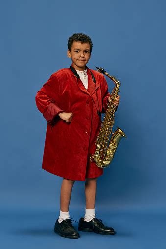 거대한 남자의 재킷과 신발을 신고 있는 귀여운 아프리카 소년의 초상화는 파란색 배경 위에 색소폰으로 연주하는 재즈 남자와 같습니다 패션 예술 음악 및 창의적인 스타일