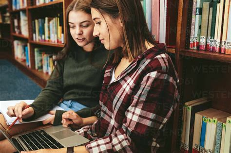 Female Students Sitting In Library With A Book And Laptop Studying