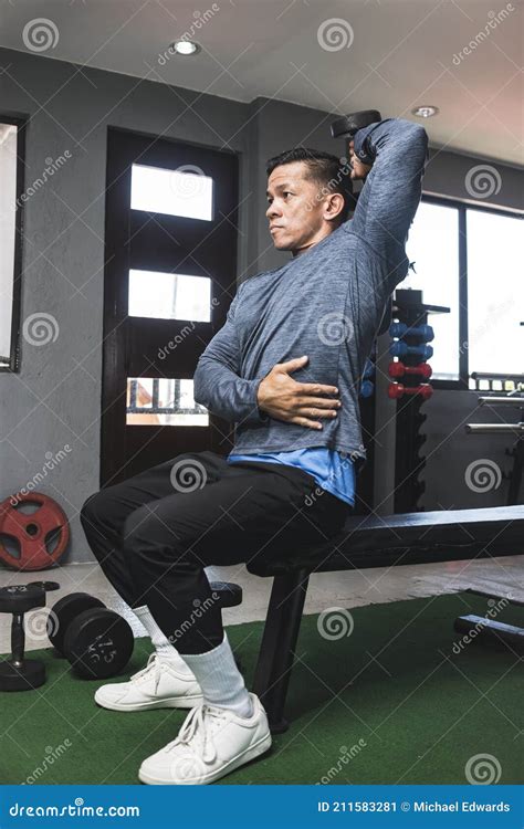 A Fit Athletic Asian Guy In A Sweatshirt Does Seated Overhead Dumbbell One Arm Tricep Extensions